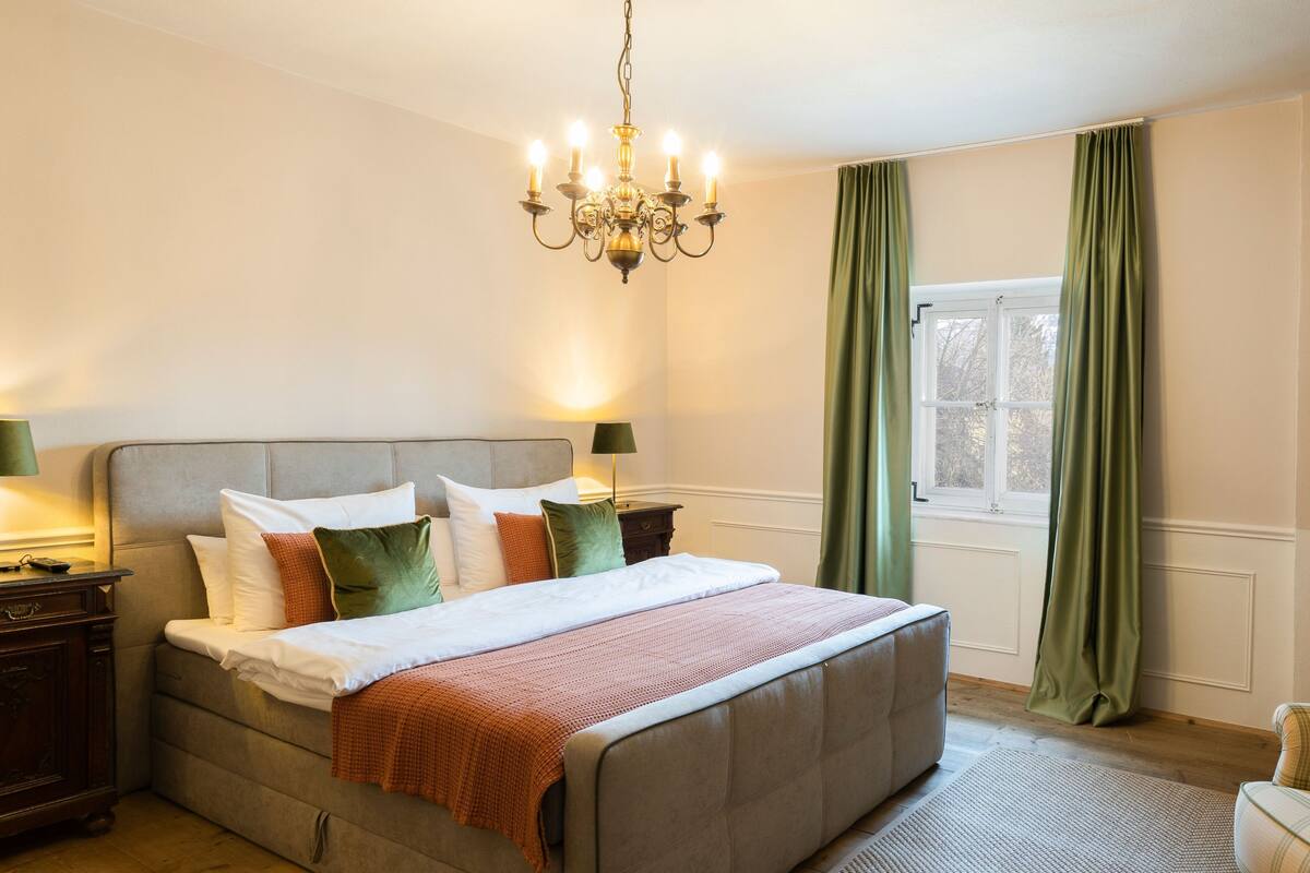 Quarto com uma cama grande, duas mesas de cabeceira, um candelabro, uma janela com cortinas e piso de madeira.