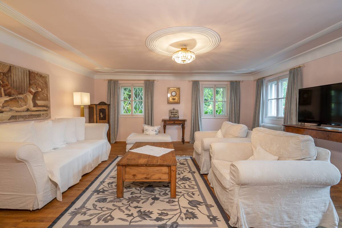 Uma sala de estar espaçosa com mesa de centro de madeira, sofás brancos, um candelabro e uma televisão.