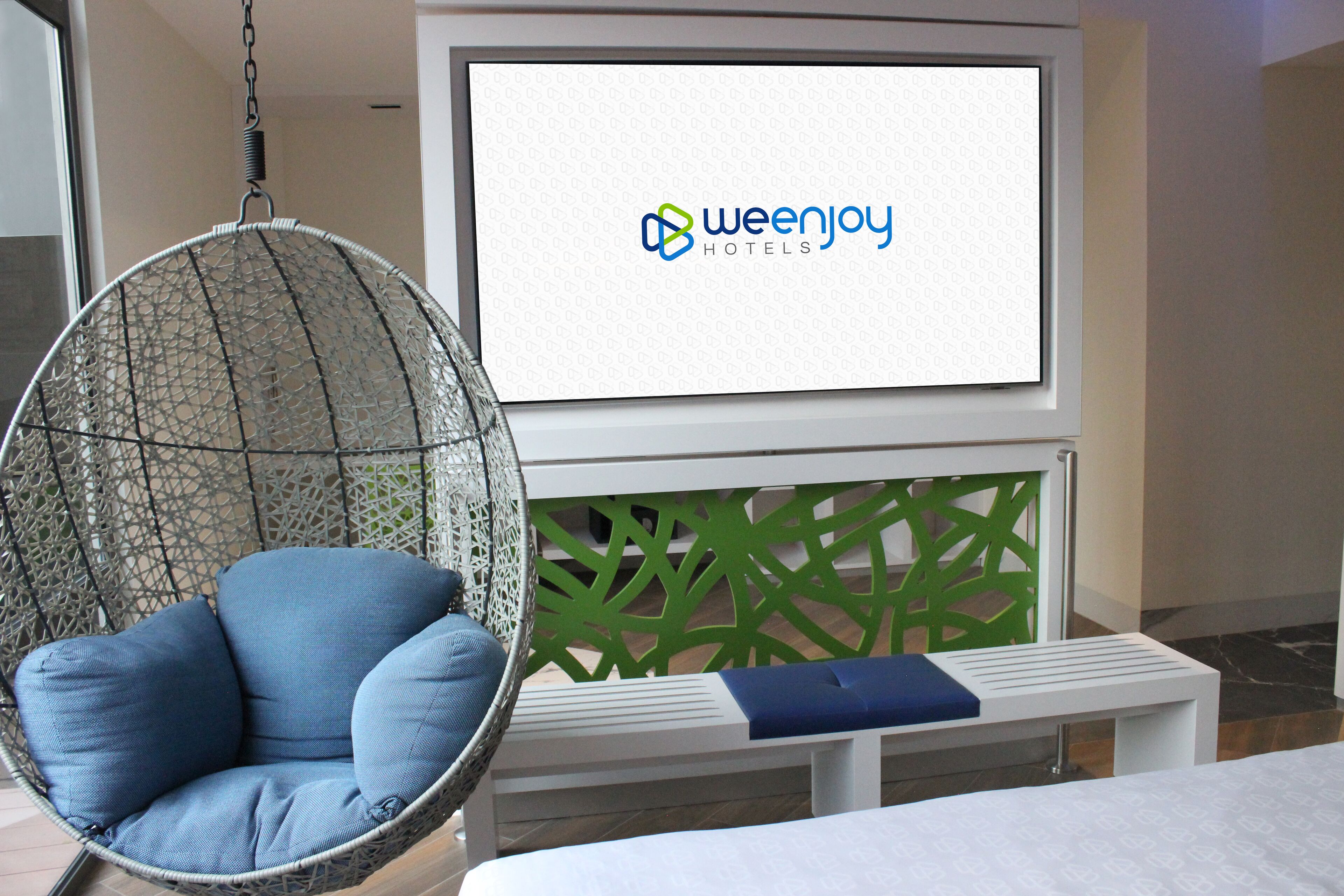 Uma cadeira suspensa com almofadas azuis, um banco branco e uma tela exibindo o logotipo Weenjoy Hotels.