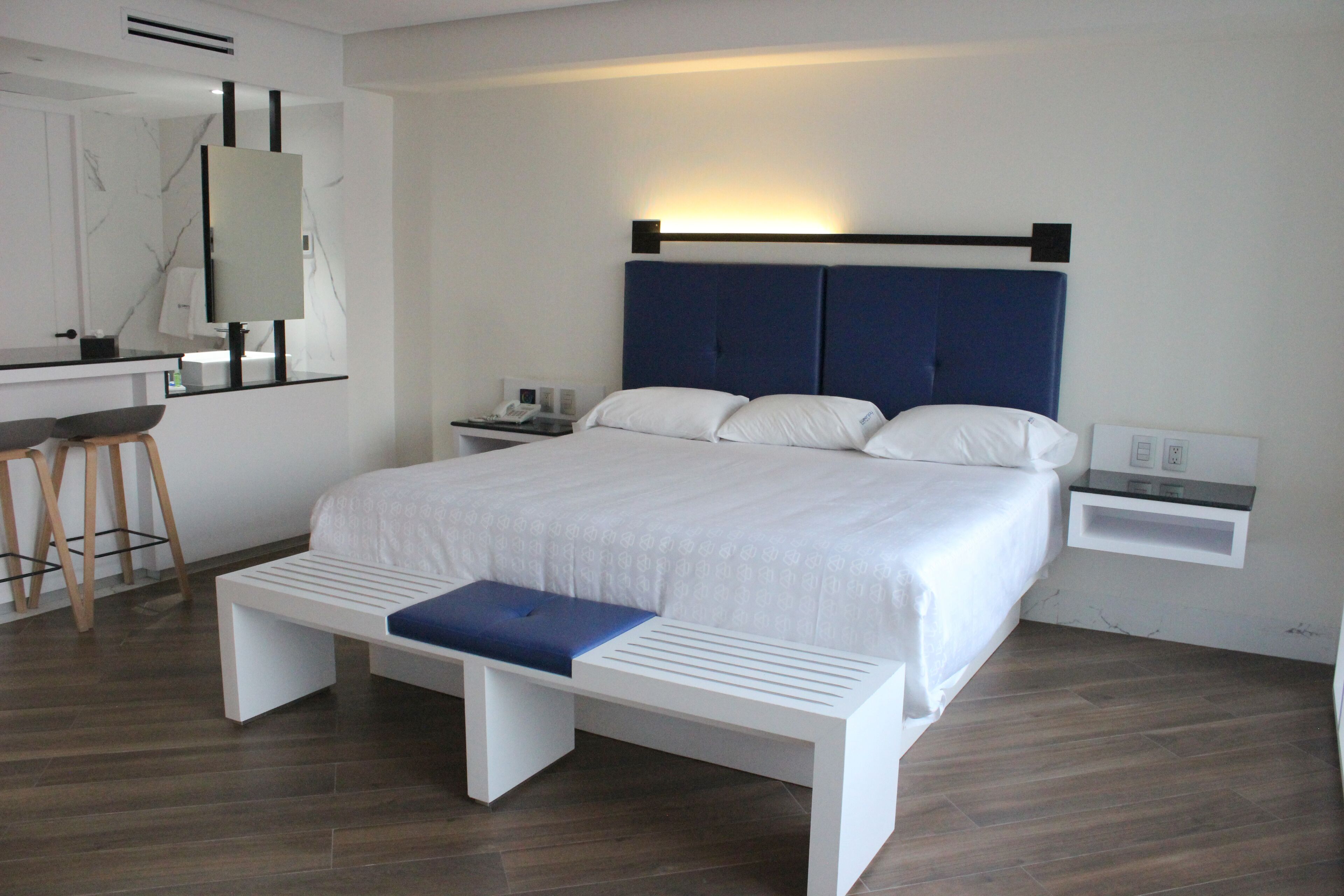 Quarto de hotel moderno com uma cama grande, uma mesa de cabeceira, uma pequena escrivaninha e uma cadeira.