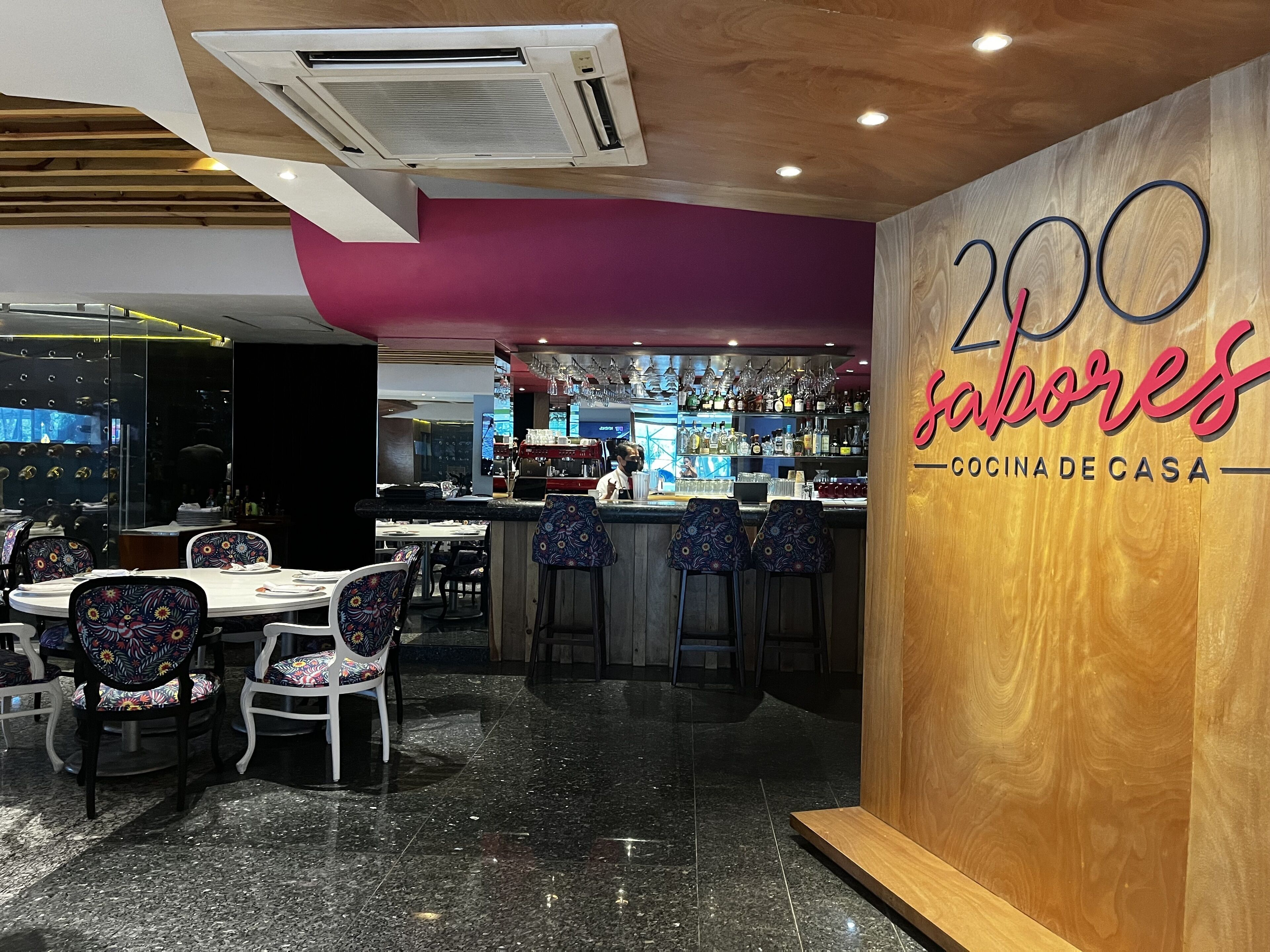 Interior de restaurante com área de bar, banquetas e um letreiro na parede que diz "200 sabores cocina de casa".