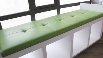 Um banco acolchoado verde com base branca, posicionado em frente a uma janela de moldura preta.