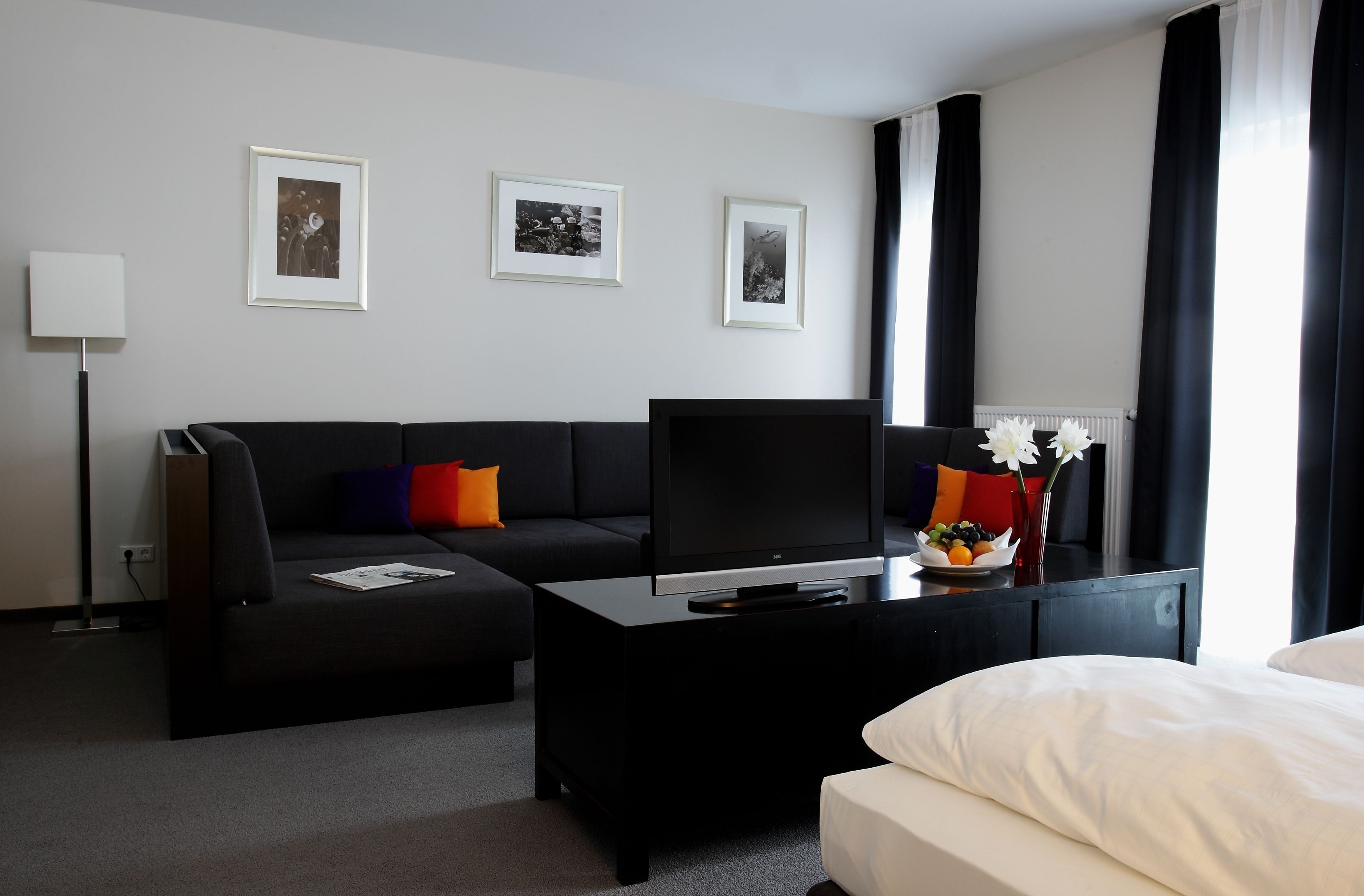 Quarto de hotel moderno com sofá preto, televisão de tela plana em suporte preto, cama branca e uma pequena mesa com um vaso de flores.
