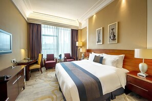Quarto de hotel com uma cama grande, uma escrivaninha, duas cadeiras, uma televisão e uma janela com cortinas.