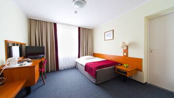 Quarto de hotel com cama, escrivaninha com computador, televisão, mesa de cabeceira com abajur e janela com cortinas.