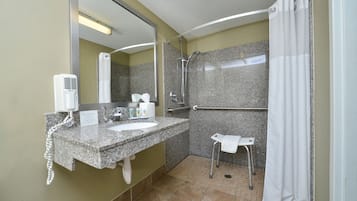 Casa de banho com bancada de granito, lavatório, espelho, secador de cabelo portátil, chuveiro com barra de apoio e toalha branca sobre um banquinho.