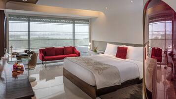 Quarto de hotel moderno com uma cama grande, um sofá vermelho, uma mesa com uma cesta de frutas e vista para a cidade através de grandes janelas.