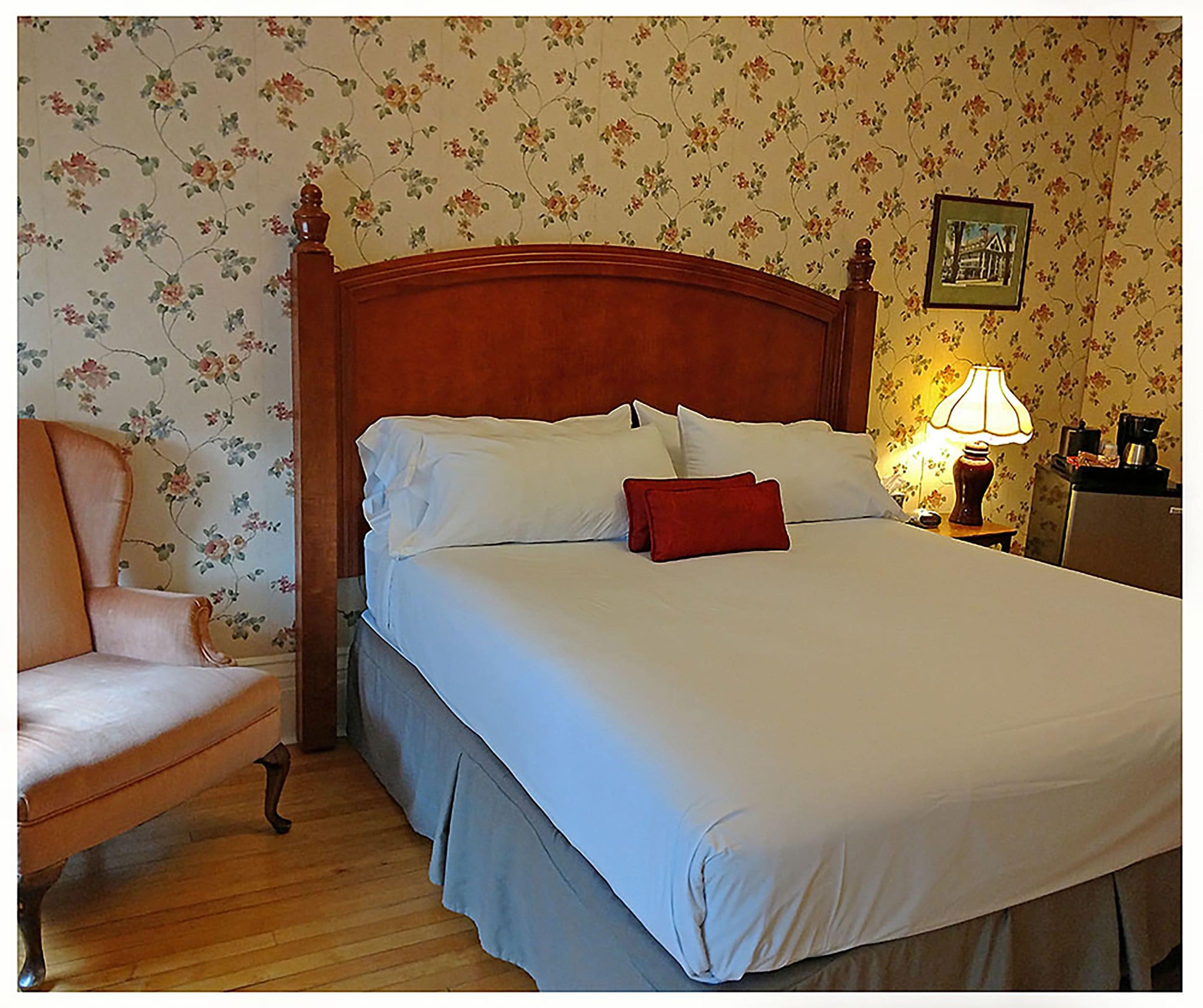 Cama bem arrumada com lençóis brancos e travesseiro vermelho, mesa de cabeceira de madeira com abajur e padrão de papel de parede floral.