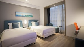 Quarto de hotel com duas camas, uma mesinha de cabeceira, uma pequena mesa, uma cadeira, um quadro e uma janela com cortinas.