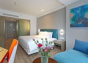 Quarto de hotel com cama, escrivaninha, cadeira, mesa de cabeceira, um vaso com flores e um quadro na parede.