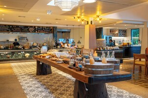 Disposição de buffet com diversos alimentos e estação de bebidas em área de refeições moderna e bem iluminada.
