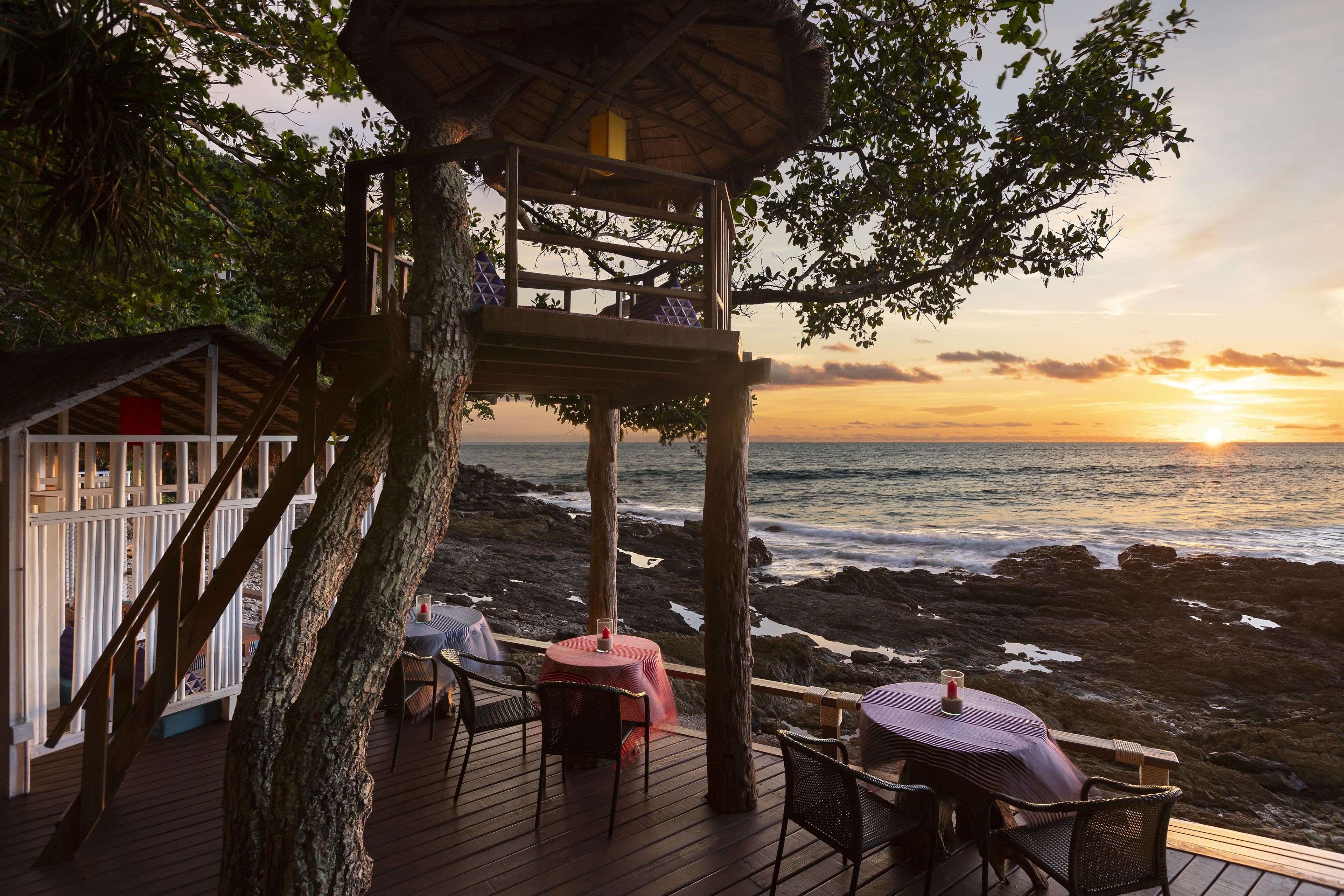 Um deck de madeira com área de estar externa com vista para uma costa rochosa ao pôr do sol.