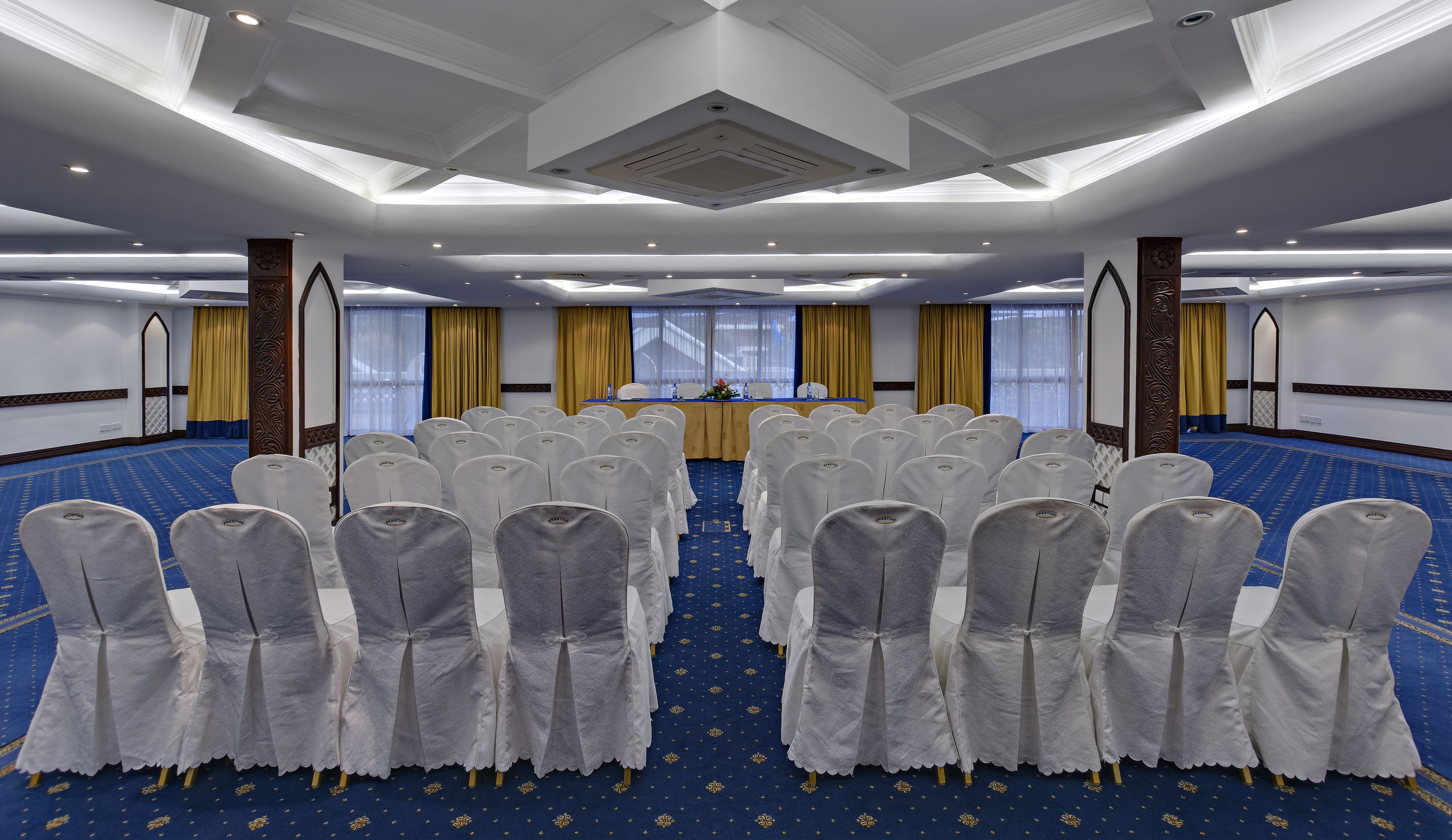 Uma sala de conferências com fileiras de cadeiras, uma mesa com um microfone e uma grande janela com cortinas.
