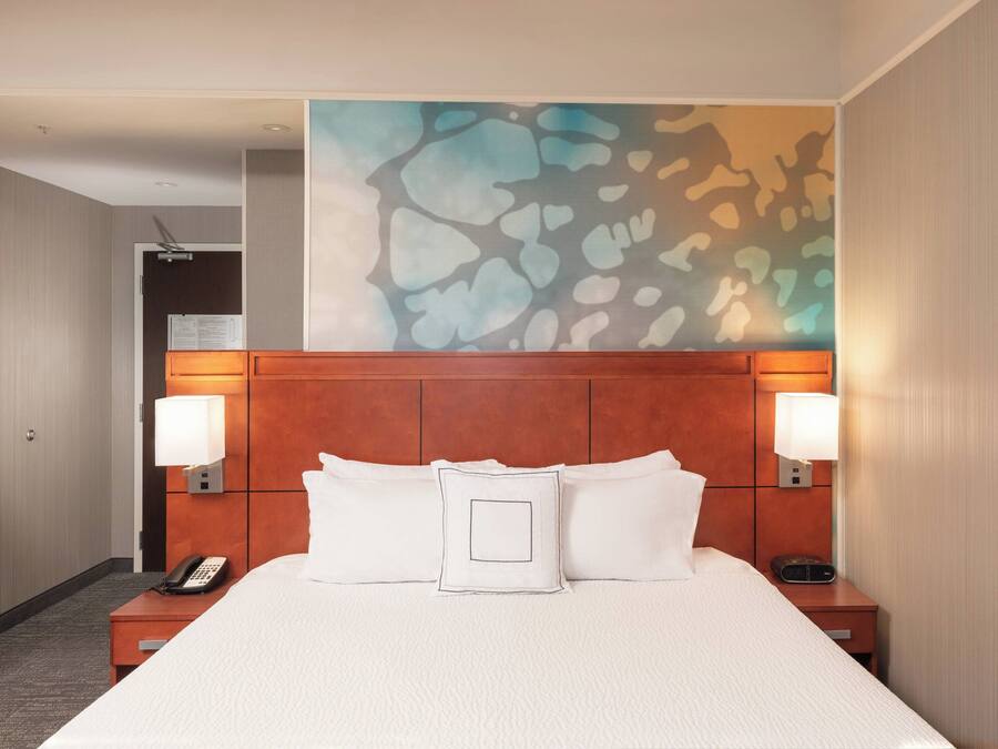 Quarto de hotel com uma cama grande, duas luminárias de cabeceira, um telefone e uma obra de arte na parede.