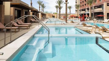 Piscina com espreguiçadeiras e palmeiras, rodeada por um edifício de hotel.
