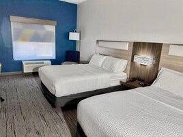 Quarto de hotel com duas camas, uma janela grande com persianas, uma mesinha de cabeceira com luminária e um aplique na parede.