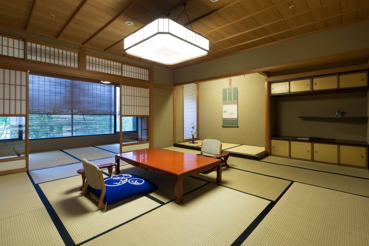 Um quarto japonês tradicional com tatames, uma mesa de madeira baixa, uma cadeira e uma janela grande com biombos shoji.