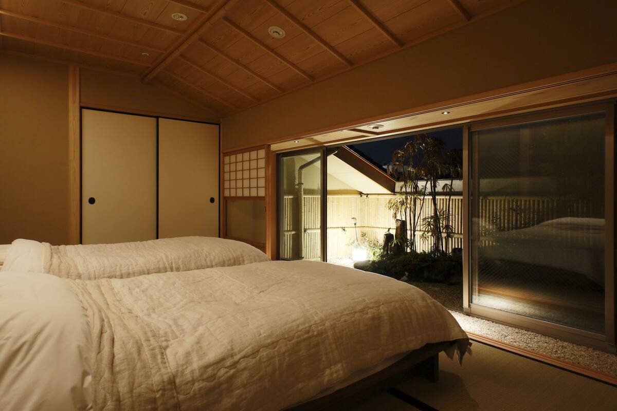 Quarto tradicional japonês com piso de tatame, porta deslizante e grande janela com vista para um jardim.