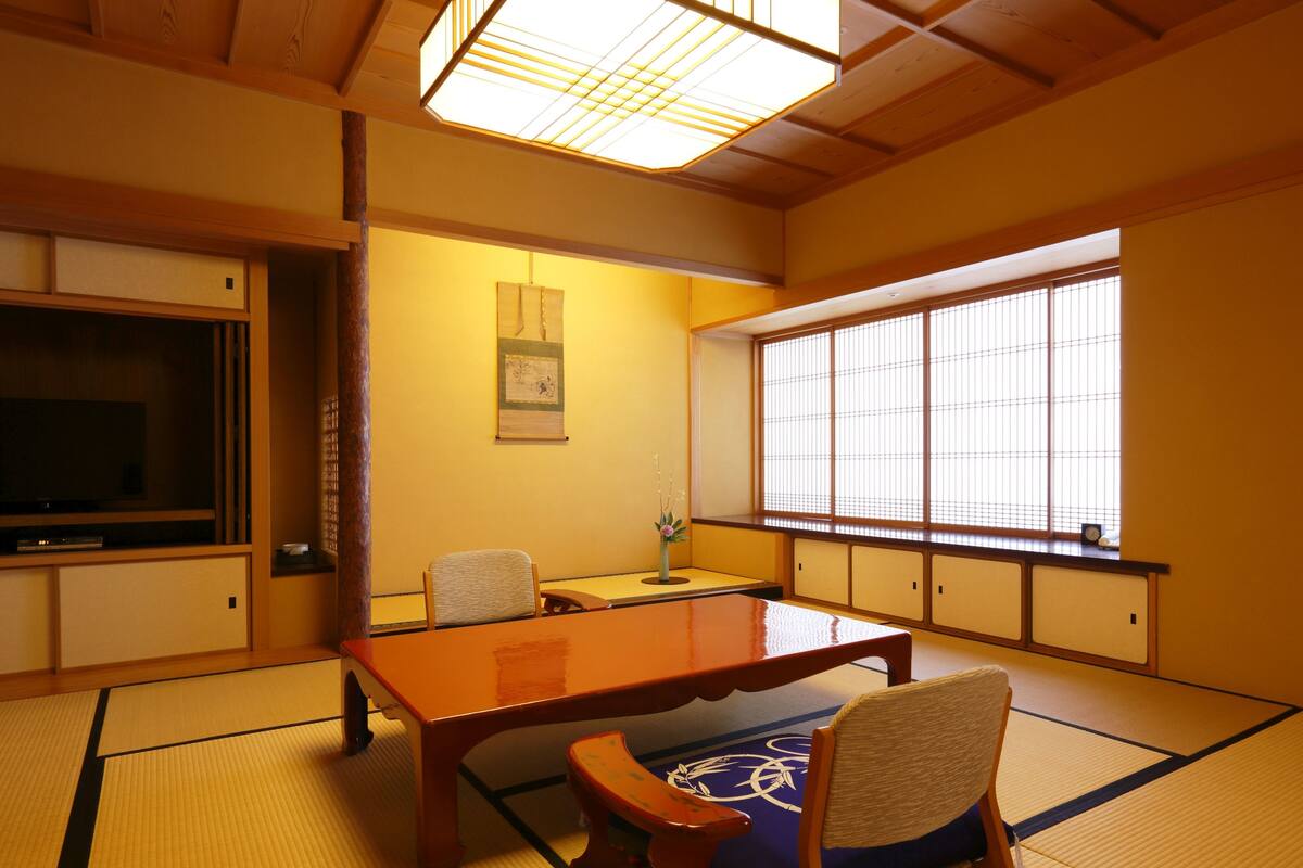 Um quarto japonês tradicional com piso de tatame, uma mesa de madeira baixa, um assento em tatame, uma televisão e uma grande janela com biombos shoji.