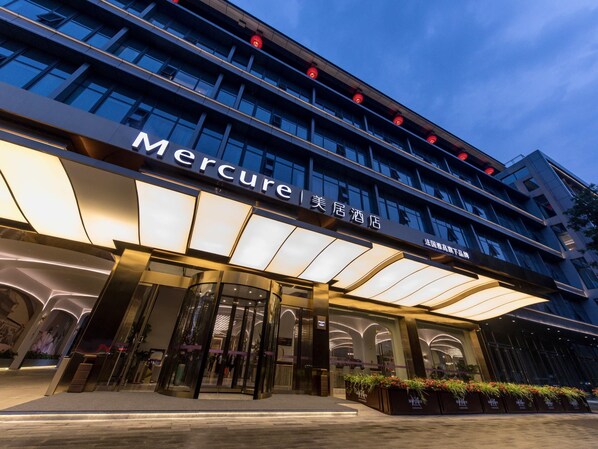 Um hotel moderno com um letreiro de 'Mercure' em destaque, grandes janelas de vidro e uma entrada bem iluminada.