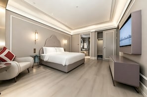 Quarto de hotel moderno com uma cama grande, área de estar com uma poltrona e uma mesa pequena, televisão fixada na parede e vista para a paisagem urbana pela janela.