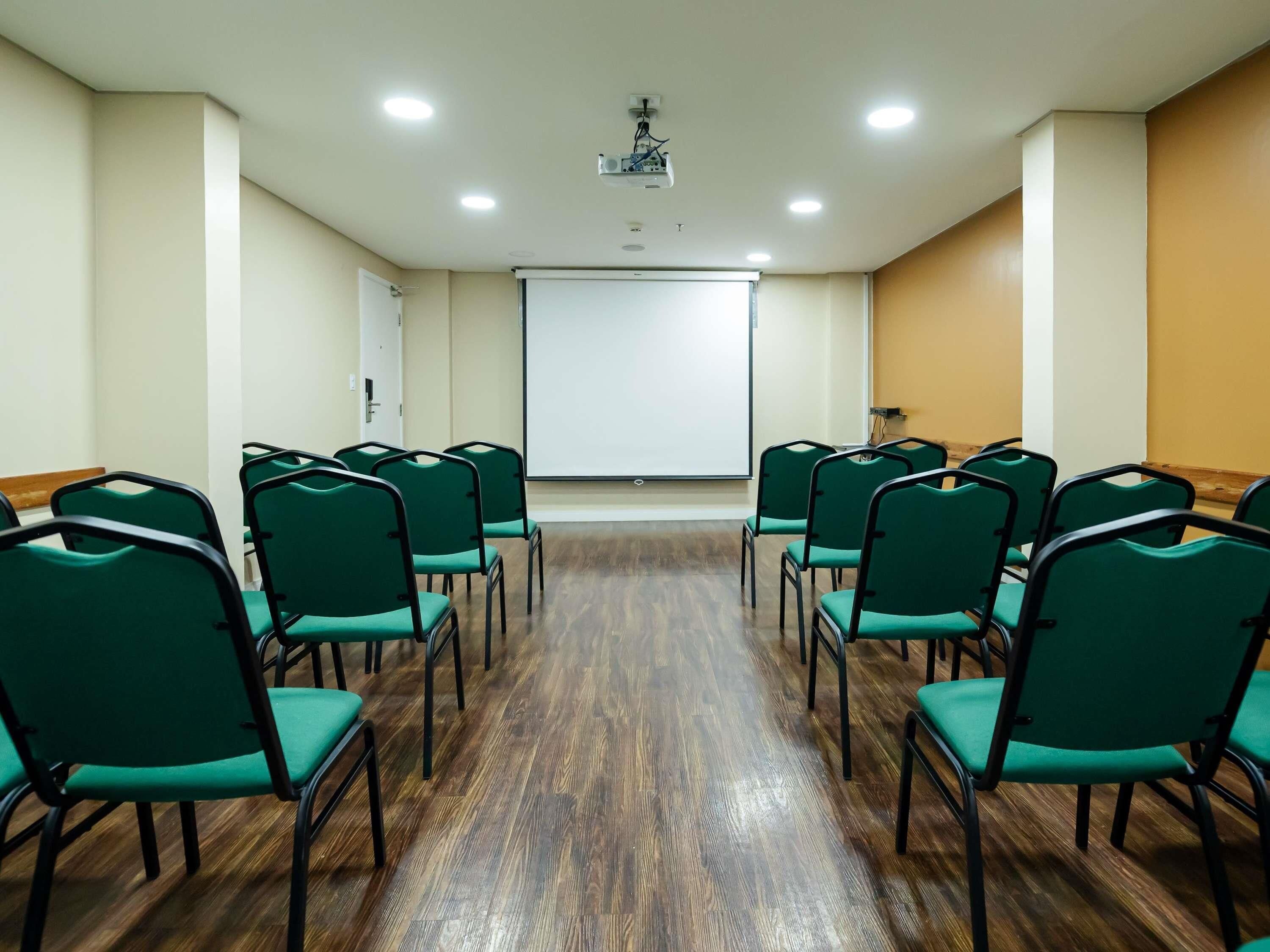 Uma sala de conferências com fileiras de cadeiras verdes, uma tela de projeção branca e piso de madeira.