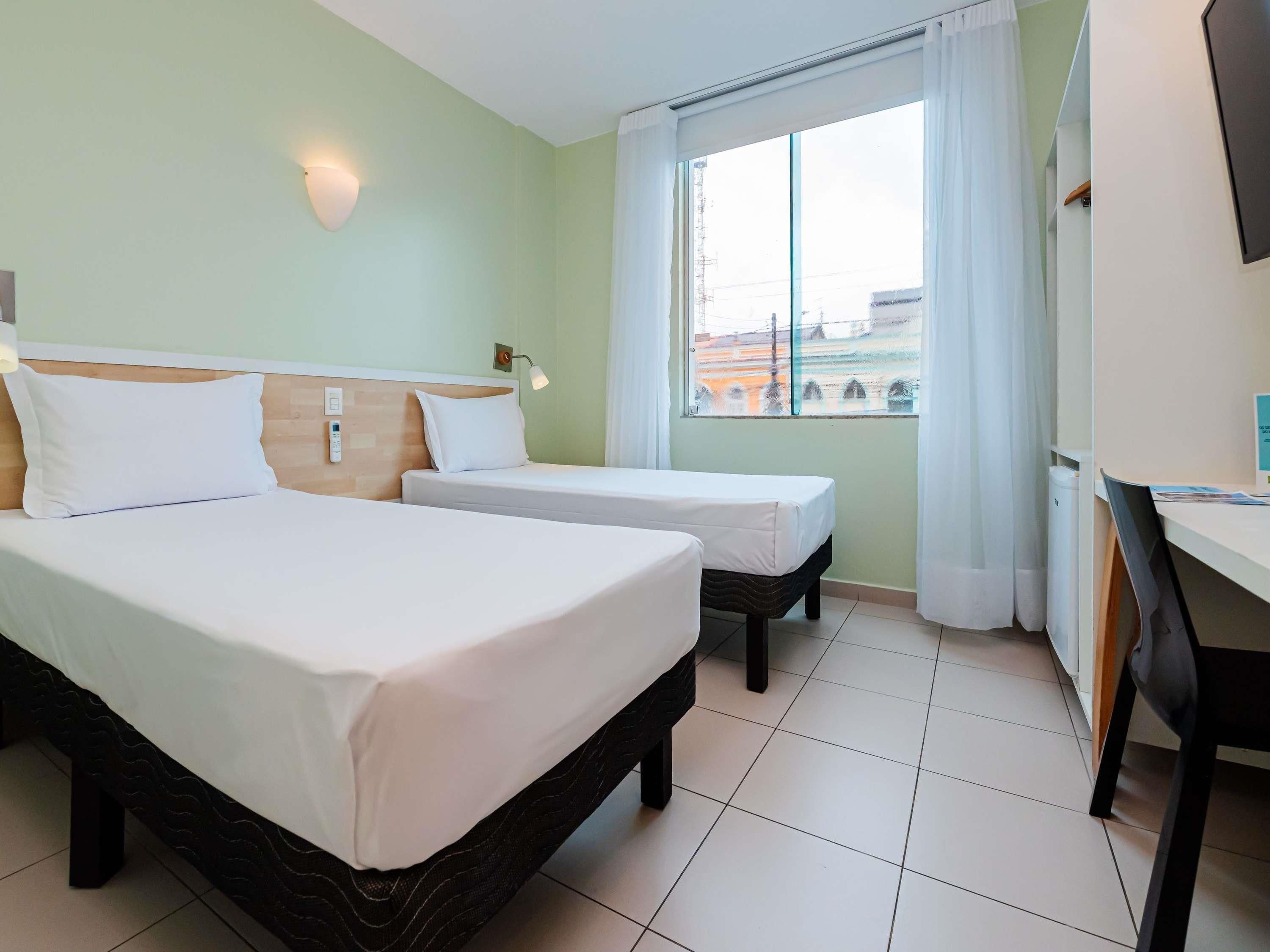 Quarto de hotel com duas camas, uma escrivaninha e uma janela com cortinas.