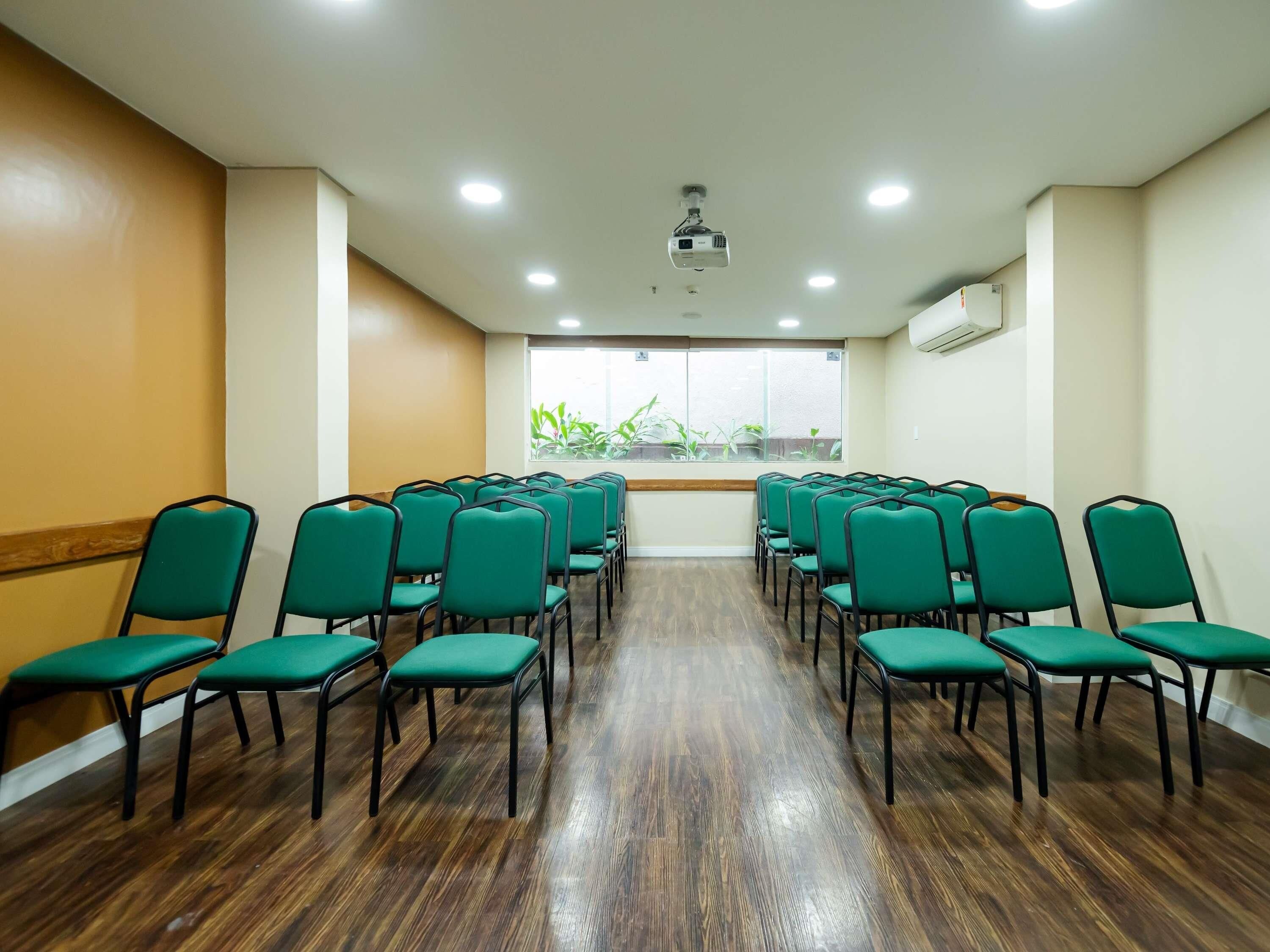 Uma sala de conferências com fileiras de cadeiras verdes, piso de madeira, um projetor e uma janela com plantas.
