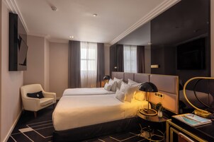 Quarto de hotel com duas camas, uma cadeira, uma escrivaninha com abajur, televisão e um espelho grande.