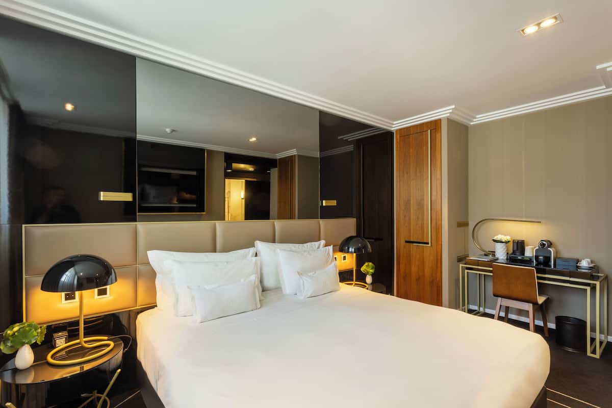Quarto de hotel com uma cama grande, abajures, uma escrivaninha e um espelho.