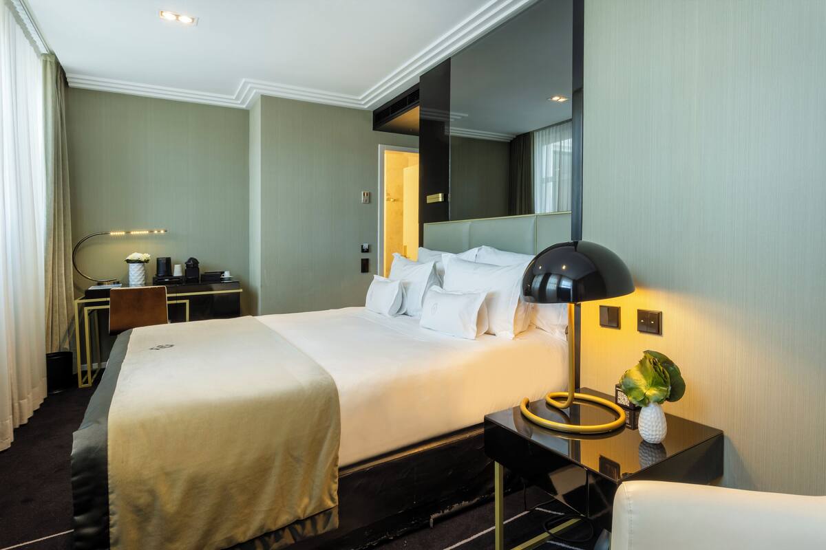 Quarto de hotel com uma cama grande, uma escrivaninha, uma cadeira, um abajur e um espelho.