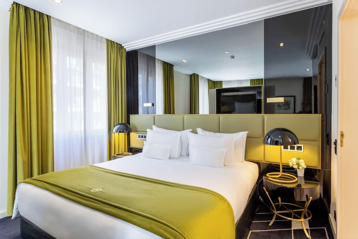 Quarto de hotel com uma cama grande, uma mesa de cabeceira com abajur, um espelho sobre a cama e vista para a cidade pela janela.