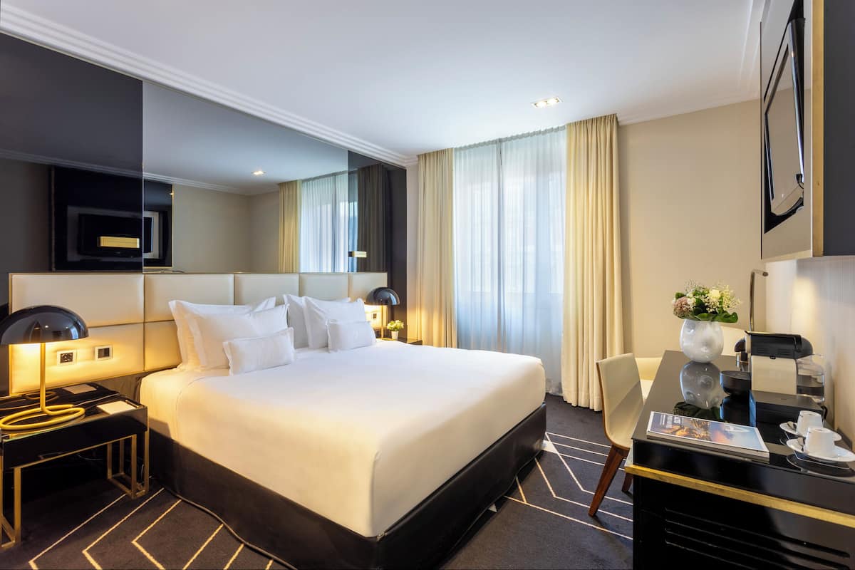 Quarto de hotel com cama grande, luminárias de cabeceira, escrivaninha com computador e um vaso com flores.