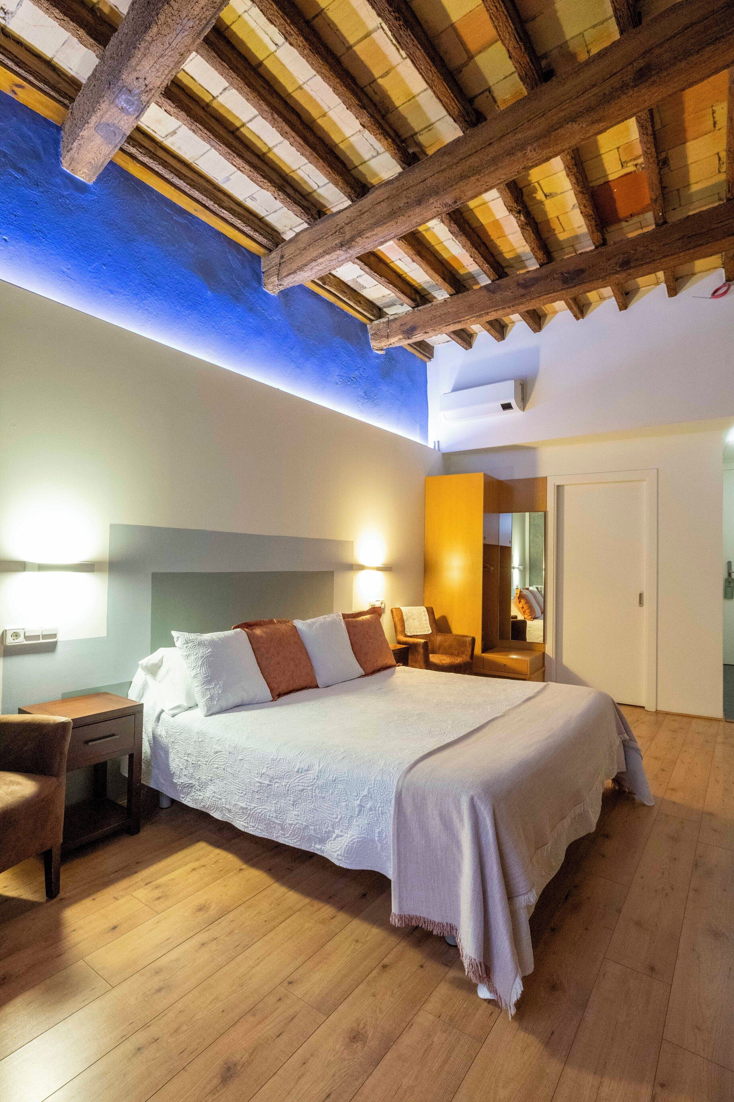 Quarto com teto de madeira, cama com roupa de cama branca, mesa de cabeceira e uma cadeira.