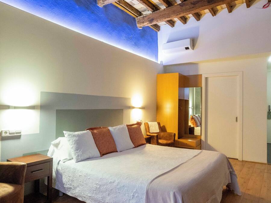Quarto com teto de madeira, cama com roupa de cama branca, mesa de cabeceira e uma cadeira.