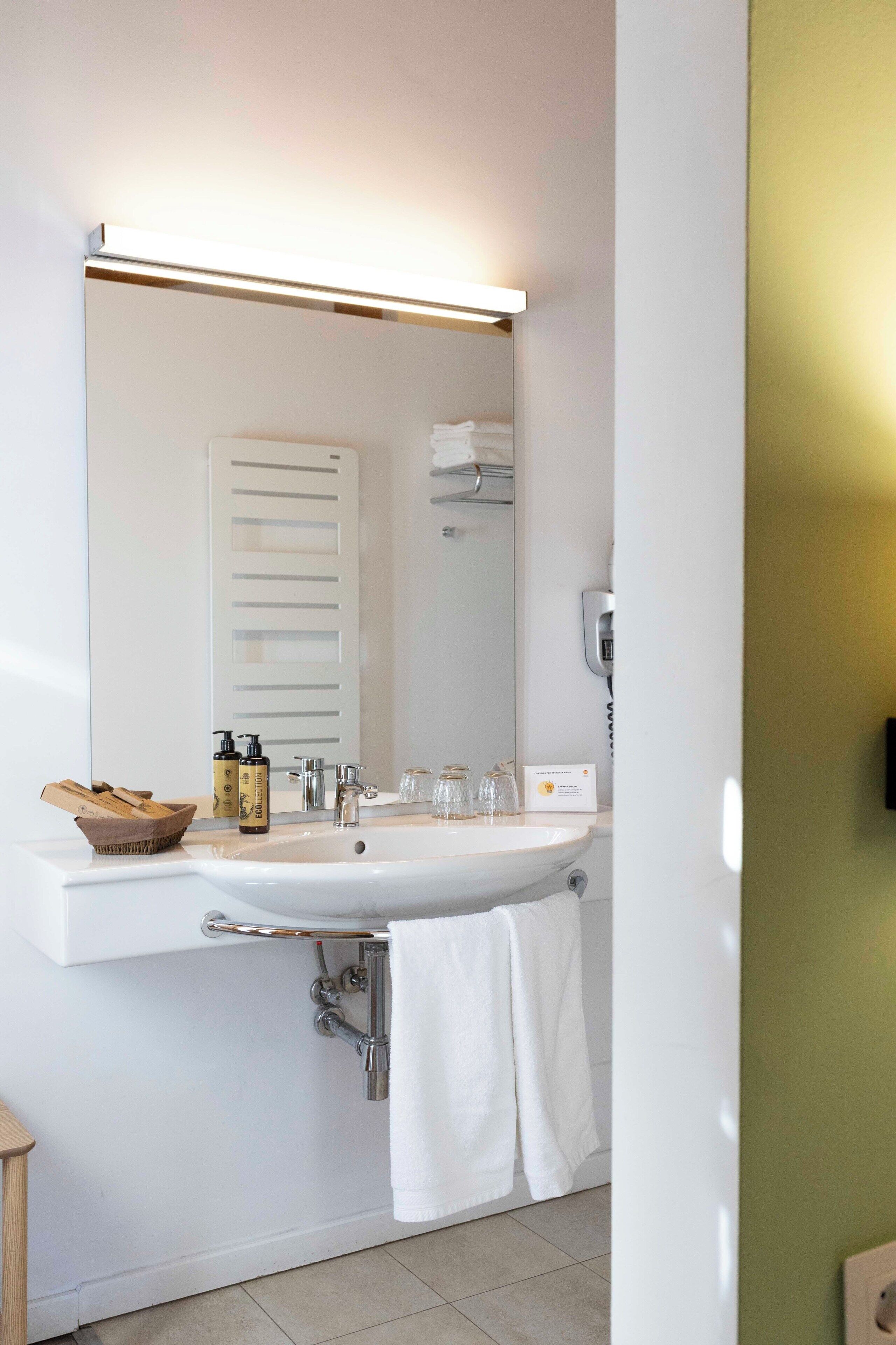 Casa de banho moderna com lavatório branco, espelho e porta-toalhas. Há produtos de higiene pessoal no lavatório e uma cesta de toalhas.