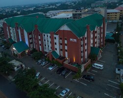 Um hotel com vários andares, telhado verde, estacionamento e edifícios ao redor.