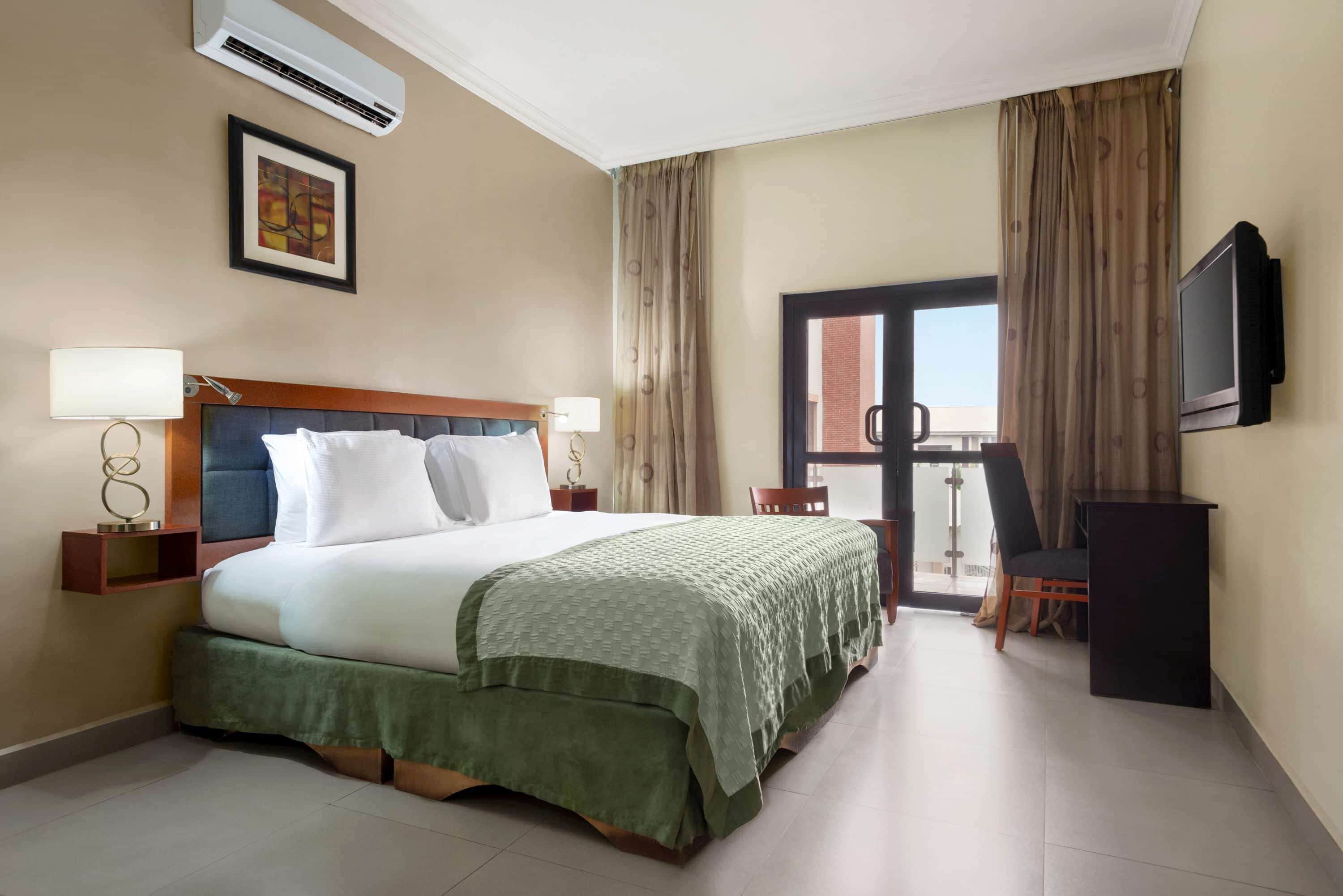 Quarto de hotel com cama, duas mesinhas de cabeceira, televisão, poltrona e varanda com cortinas.