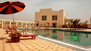 Uma área de piscina com espreguiçadeiras e guarda-sóis, um edifício com grandes janelas e um céu claro.