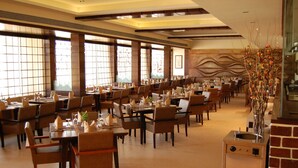 Uma sala de jantar espaçosa com várias mesas e cadeiras, janelas amplas e um painel decorativo.