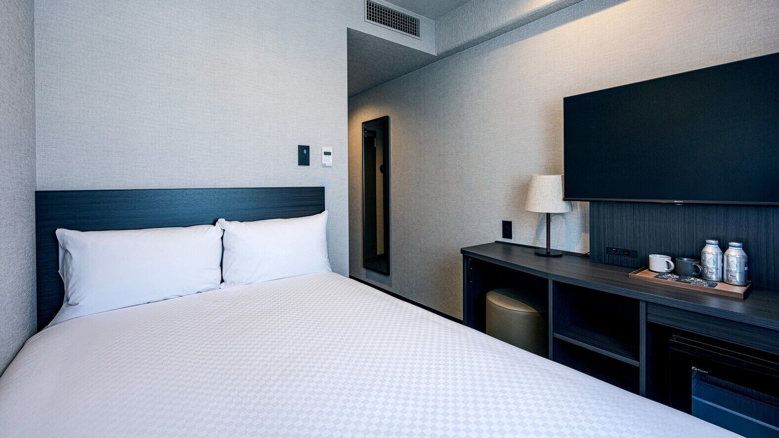 Quarto de hotel com cama, escrivaninha com luminária, TV de tela plana e minibar.