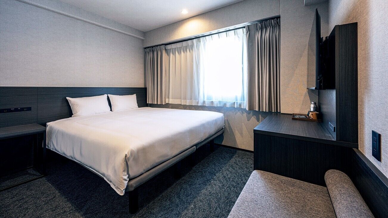 Quarto de hotel com cama, mesa de cabeceira, área de estar, televisão e janela com cortinas.