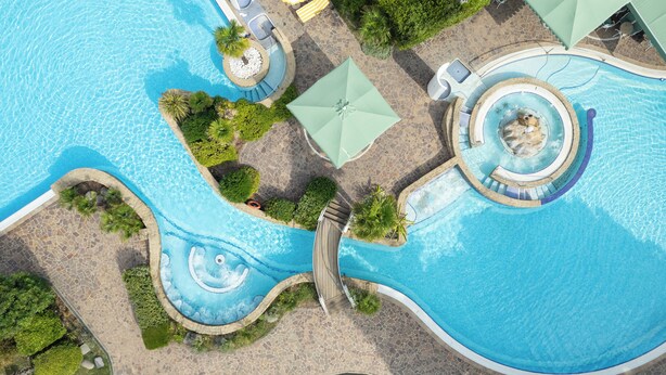 Uma piscina grande e sinuosa com um recurso aquático central, rodeada por espreguiçadeiras e guarda-sóis.