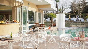 Área de piscina ao ar livre com cadeiras e mesas de metal branco, um bar com uma cesta de frutas e garrafas, e vista para a piscina e espreguiçadeiras.