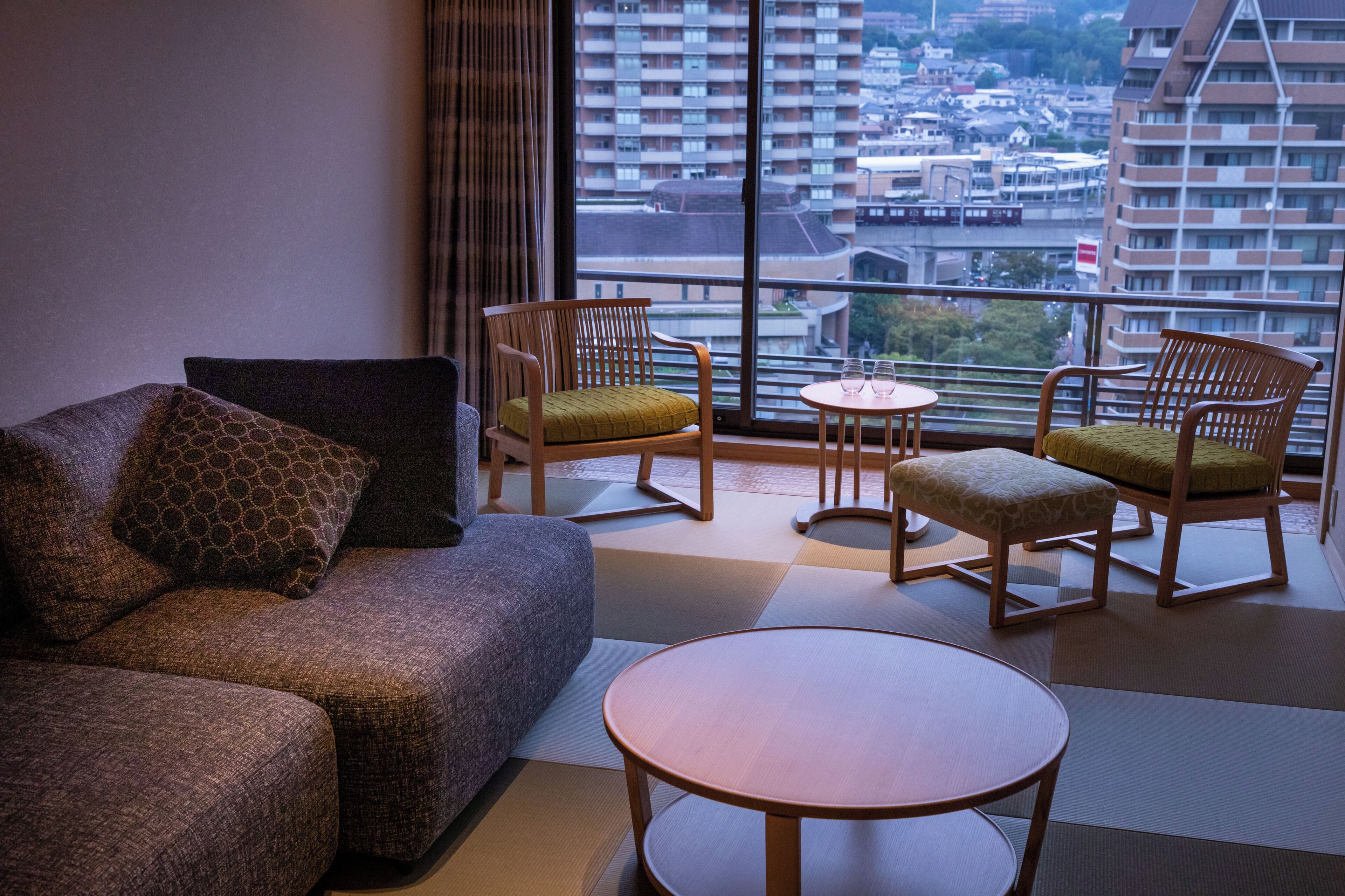 Sala de estar moderna com sofá, duas poltronas, uma mesa pequena e vista para o horizonte urbano através de grandes janelas.