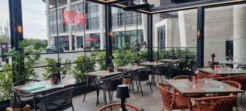 Uma área de refeições ao ar livre com mesas e cadeiras, um letreiro de neon para um restaurante e vista para a rua.