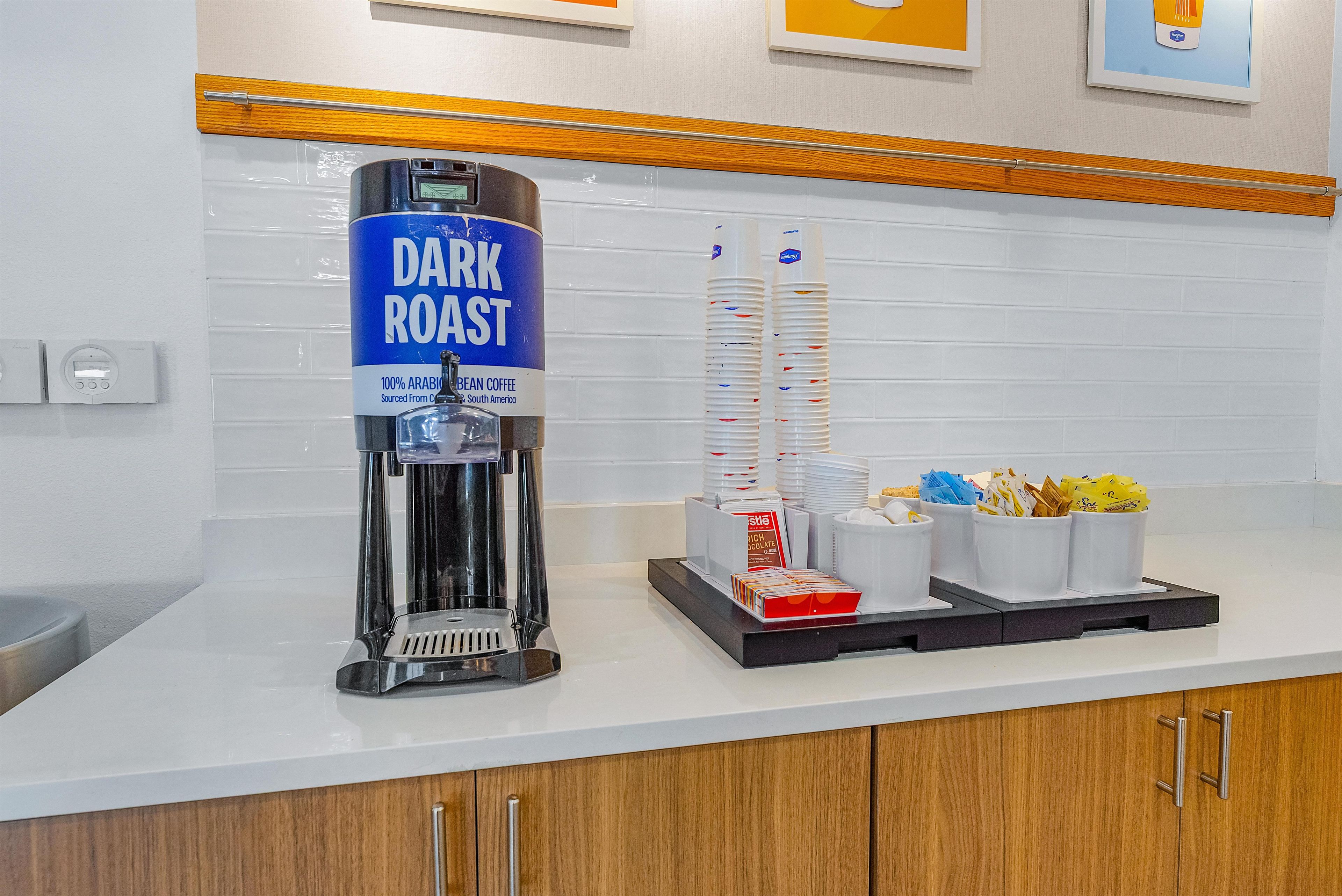 Estação de café com máquina de café rotulada 'DARK ROAST' e uma pilha de copos descartáveis.