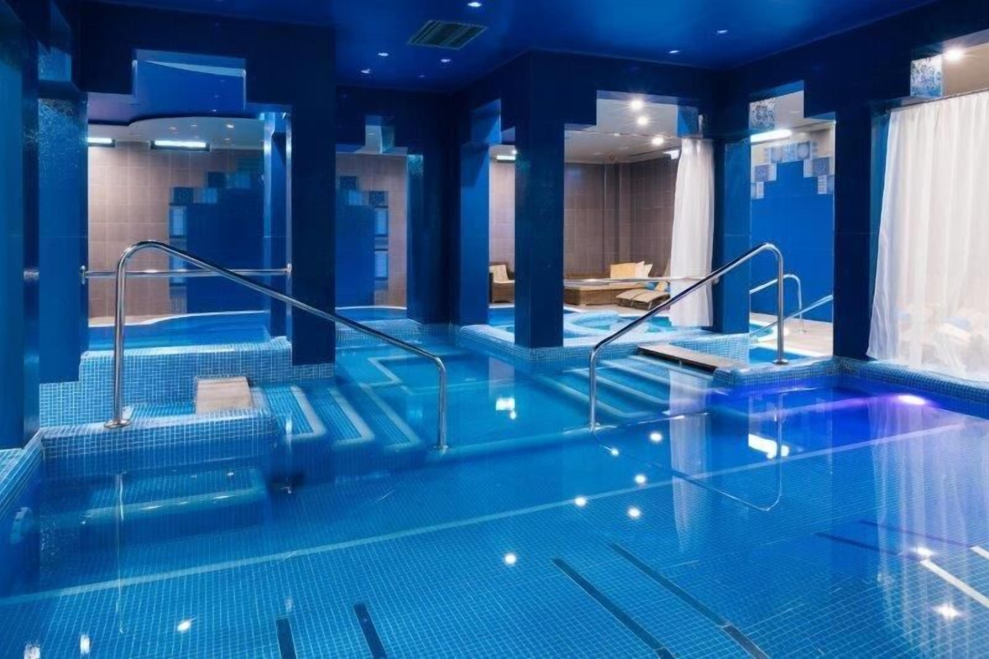 Uma grande piscina interior com água cristalina, rodeada por paredes azuis e degraus revestidos de azulejos.