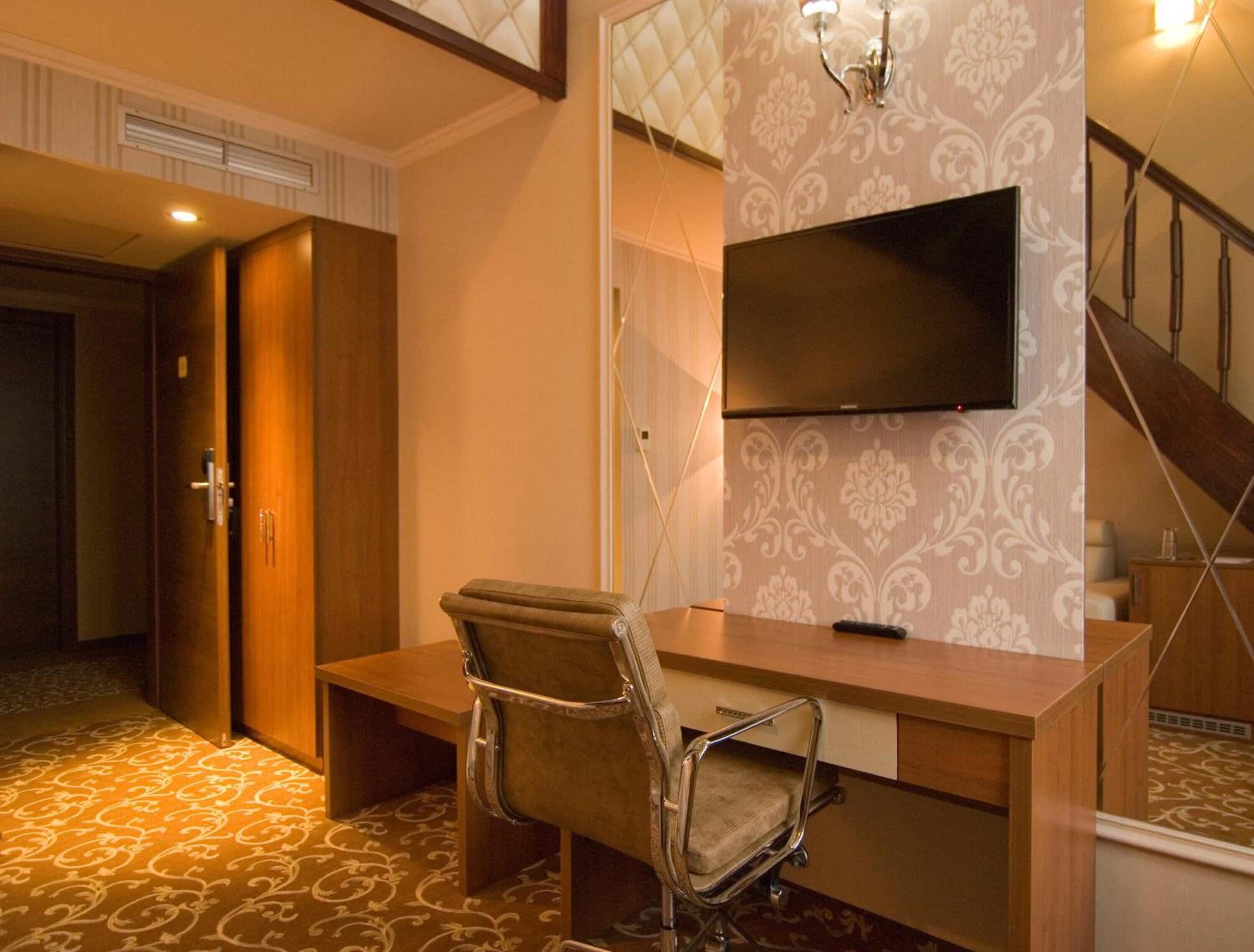 Quarto de hotel com uma escrivaninha de madeira, uma cadeira, uma televisão fixada na parede e uma escada ao fundo.