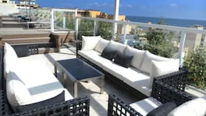 Área externa com assentos, incluindo um sofá branco, cadeiras pretas e uma mesa. Há guarda-sóis e vista para o mar.